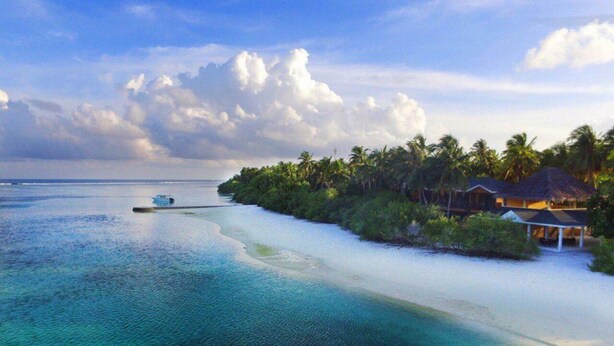 Uma praia tropical com águas turquesa cristalinas, um pequeno barco e um resort com construções de telhado de palha.