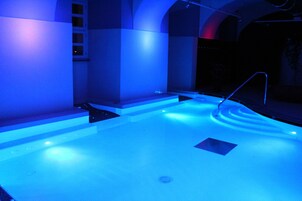 Uma piscina interior iluminada em azul, com escadas que levam à água, cercada por paredes e teto com detalhes arquitetônicos.