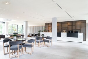 Um hall de hotel moderno com um balcão de recepção de mármore, cadeiras estofadas em cinza, uma mesa de jantar redonda e grandes janelas com vista para o verde lá fora.