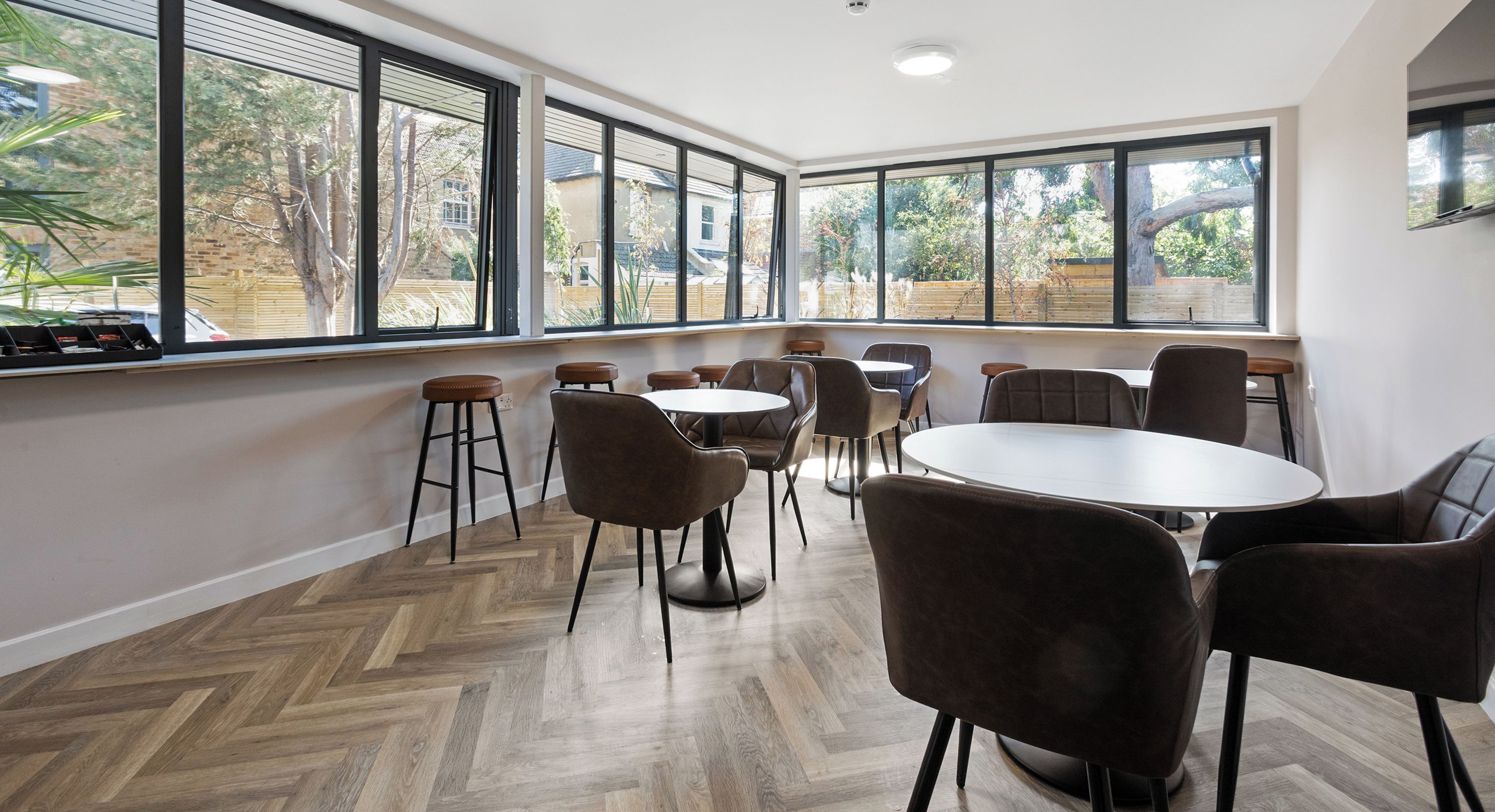 Um espaço interior moderno com janelas amplas, piso de madeira e uma mistura de mesas redondas e quadradas com cadeiras.