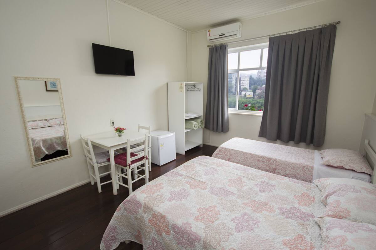 Quarto de hotel com duas camas, uma escrivaninha, uma cadeira, um frigorífico pequeno, uma televisão fixada na parede e uma janela com cortinas.
