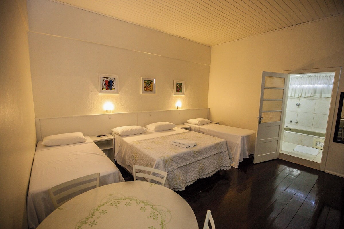 Quarto de hotel com três camas de solteiro, uma mesinha e área de chuveiro.