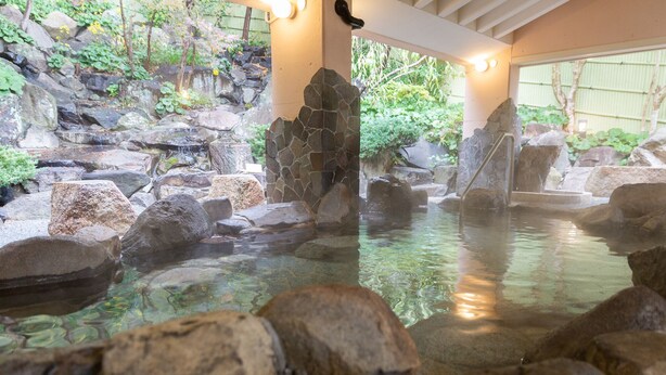 Uma área de águas termais com grandes rochas e uma piscina cristalina, cercada por paredes de pedra e vegetação.