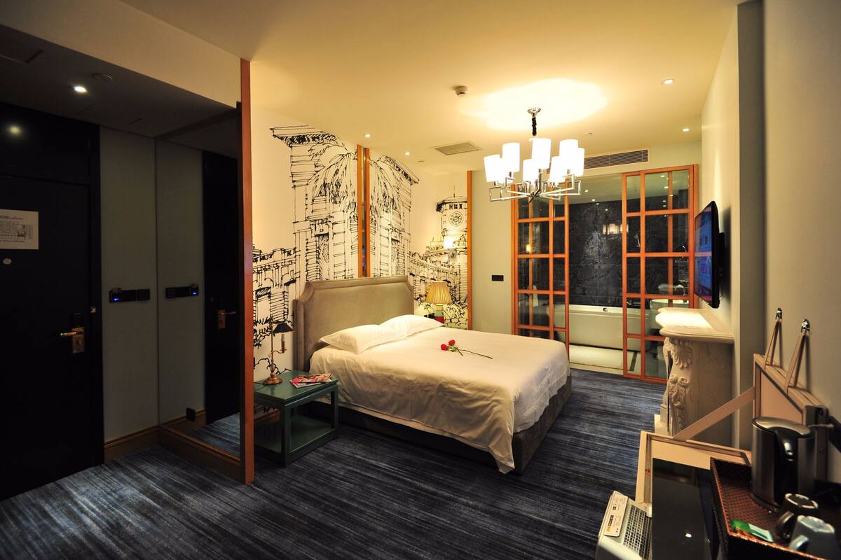 Quarto de hotel com uma cama, escrivaninha, cadeira, luminária, televisão e uma janela grande.