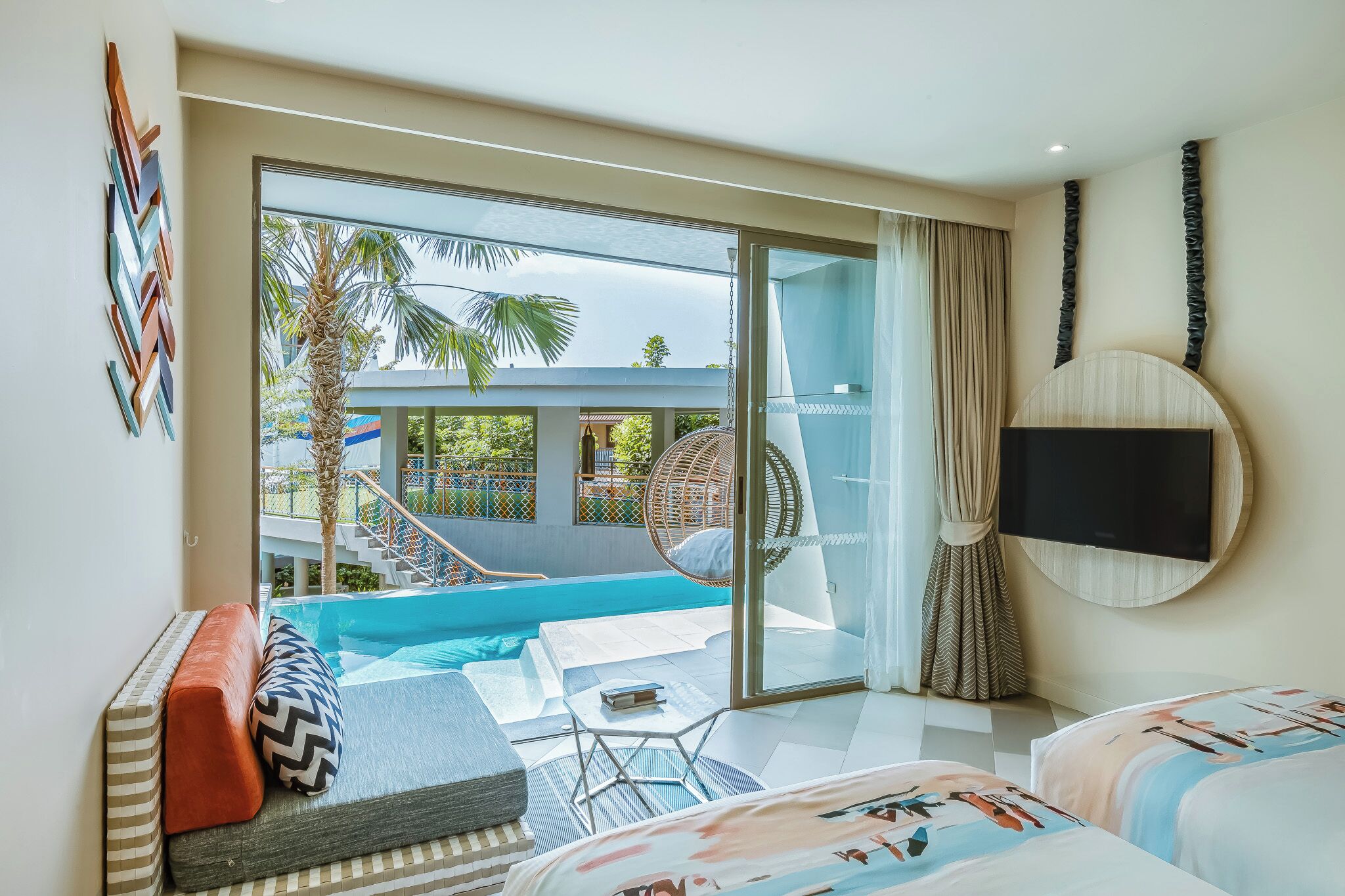 Quarto de hotel moderno com uma grande porta de vidro deslizante para uma área de piscina, uma TV de tela plana na parede e um espaço de estar confortável com almofadas.