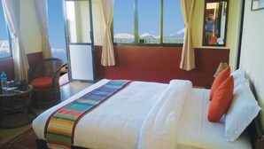 Quarto de hotel com cama, uma cadeira, uma mesa e vista para montanhas através de janelas amplas.