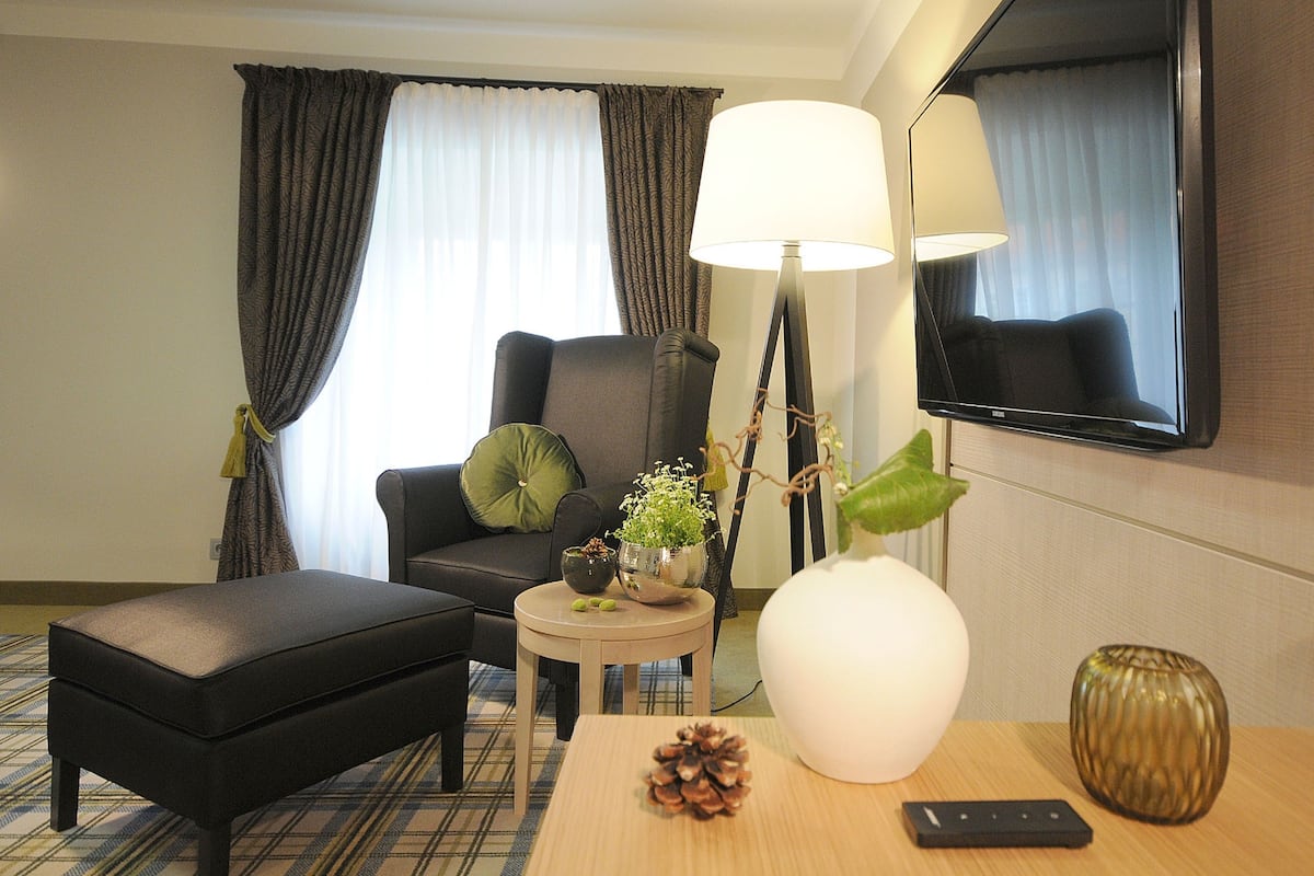 Quarto de hotel com sofá, uma pequena mesa redonda com abajur, um vaso grande e televisão na parede.