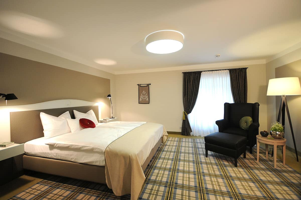 Quarto de hotel com cama, uma cadeira, uma mesa pequena e um abajur.