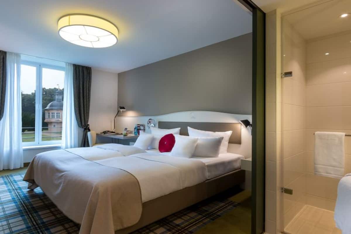 Quarto de hotel com uma cama grande, escrivaninha, cadeira, janela com cortinas e banheiro.