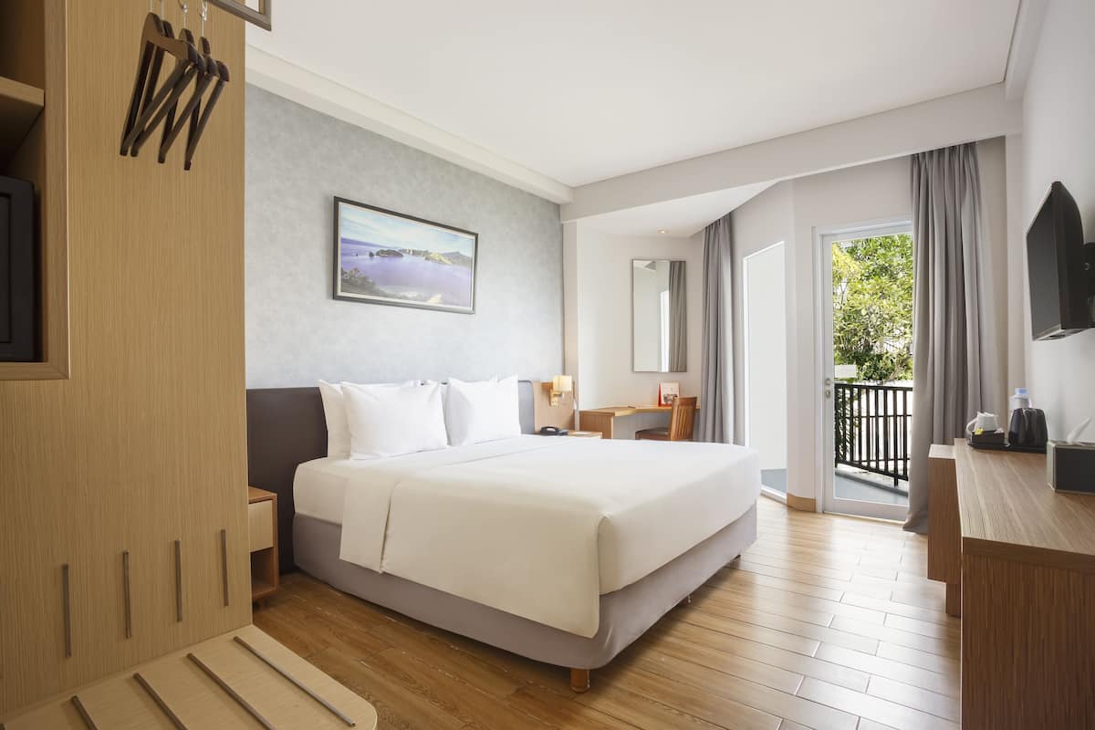 Quarto de hotel com cama grande, guarda-roupa de madeira, escrivaninha com cadeira, televisão e varanda com vista para o verde.