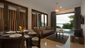 Uma sala de estar moderna com um sofá, uma mesa de jantar posta para dois e uma pequena mesa redonda com frutas. Há uma televisão em um móvel e uma janela grande com vista para o exterior.