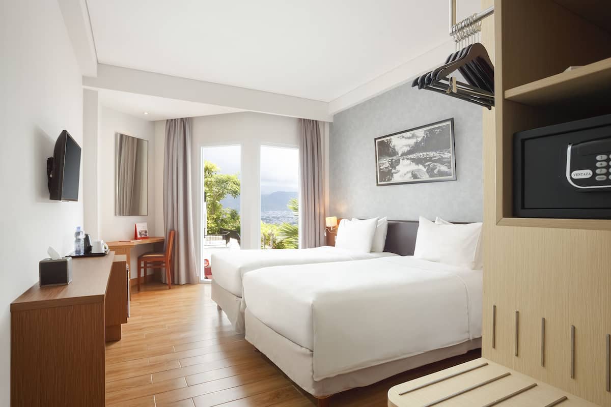Quarto de hotel com duas camas, uma escrivaninha, televisão e vista para o exterior.