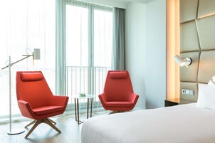 Quarto de hotel com duas cadeiras vermelhas, uma cama, uma mesa de cabeceira, um abajur e uma janela grande com cortinas.