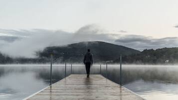 Uma pessoa caminhando em um cais de madeira que se estende por um lago calmo, cercado por névoa e montanhas.