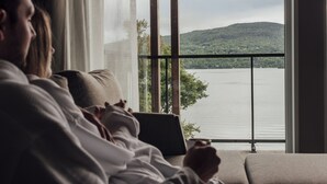 Quarto com uma grande janela com vista para um lago e colinas. Há um sofá com almofadas e uma pessoa sentada nele.