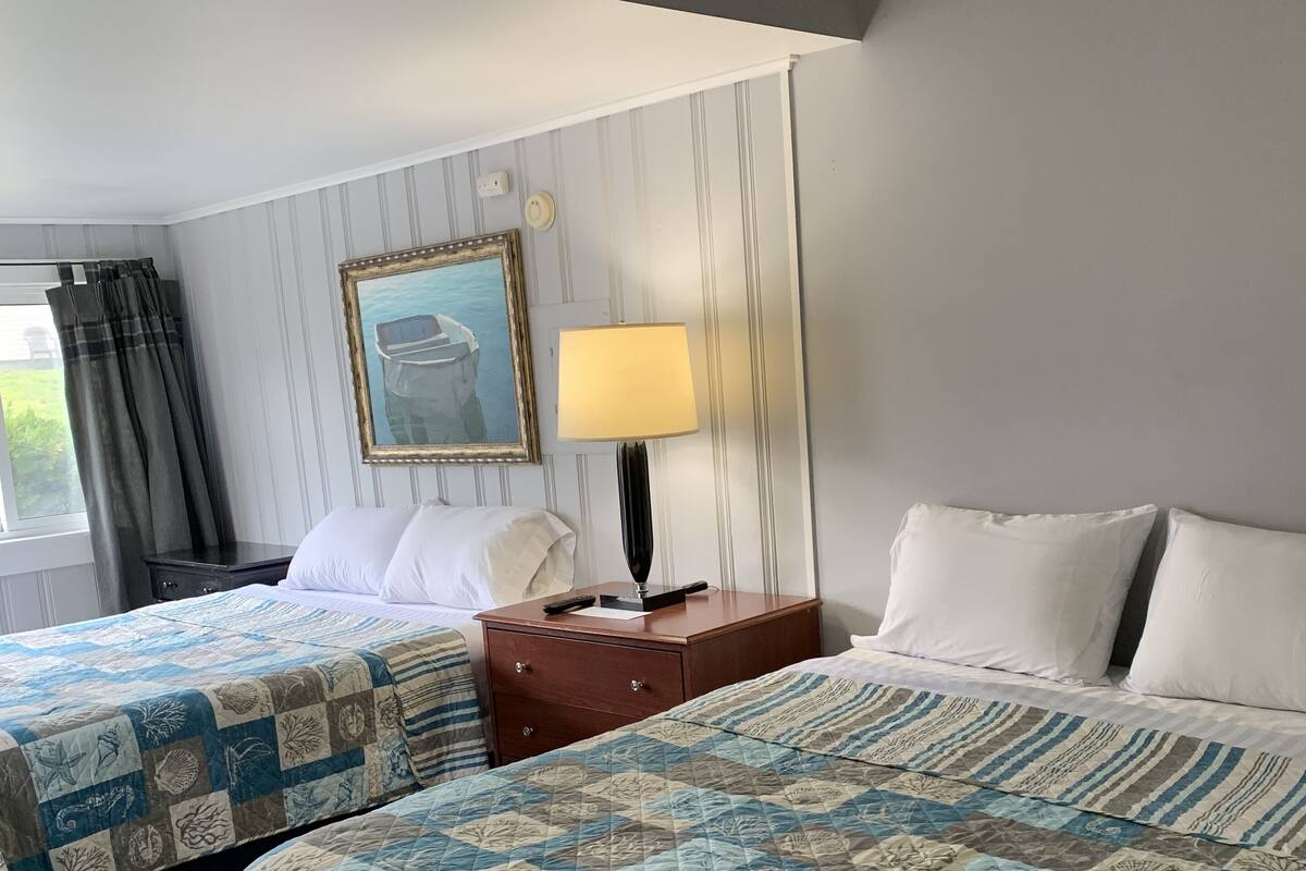 Quarto de hotel com duas camas, uma mesinha de cabeceira, uma luminária, um quadro e uma janela com cortinas.