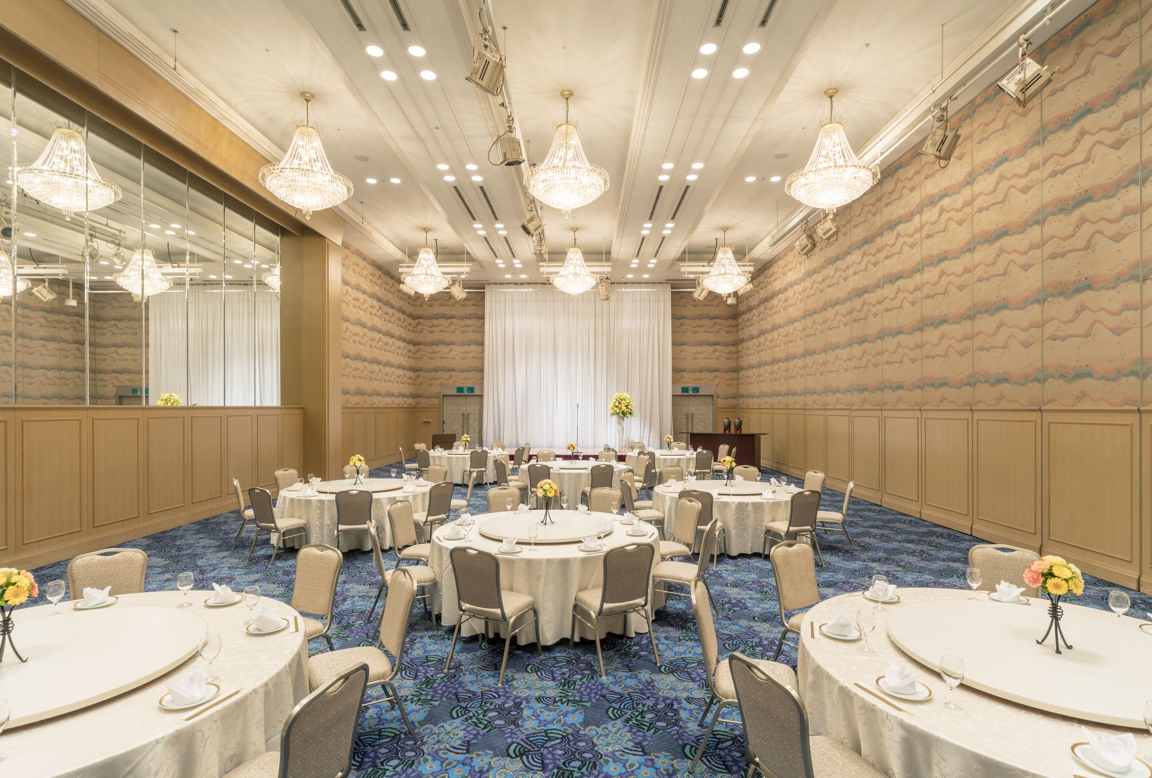 Salão de banquetes com mesas redondas, cadeiras, lustres e um tapete estampado.
