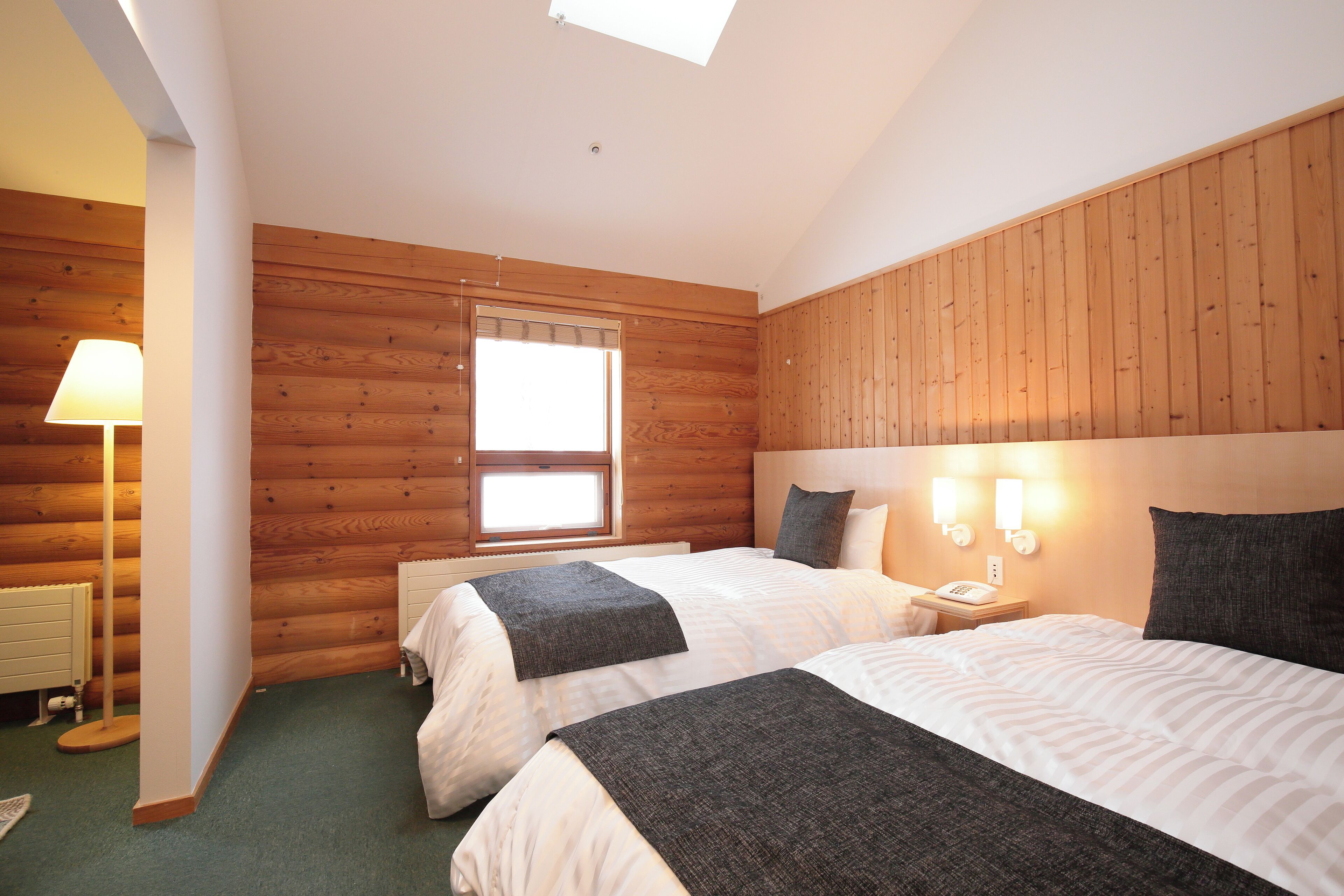 Quarto com duas camas, paredes de madeira, uma janela, um abajur e um telefone.