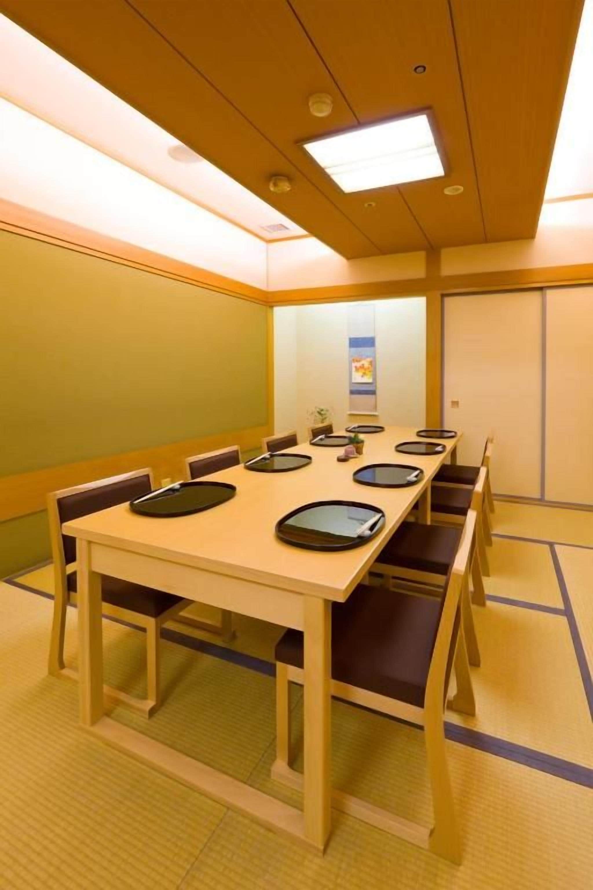Uma sala de conferências com uma mesa comprida e cadeiras, uma luminária no teto e uma porta ao fundo.