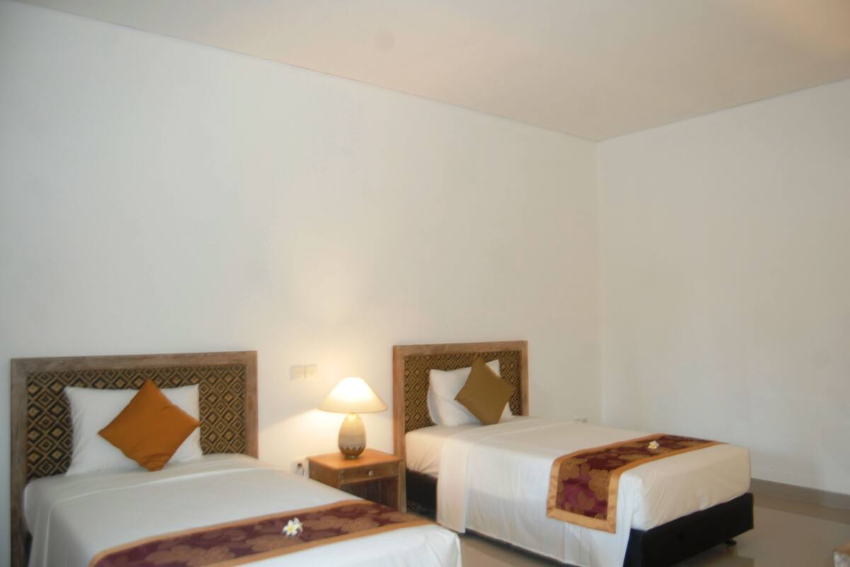 Quarto de hotel com duas camas de solteiro, cada uma com cabeceira estampada, mesa de cabeceira com abajur e decoração floral na cama.