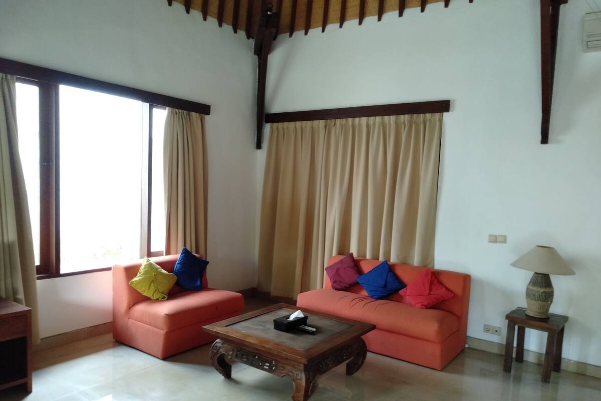 Sala de estar com dois sofás, uma mesa de centro, um abajur e janelas amplas com cortinas.