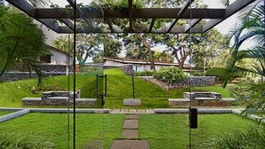 Uma área de jardim envidraçada com um caminho de pedra que leva a um espaço de estar, cercada por vegetação exuberante e um muro de pedra.