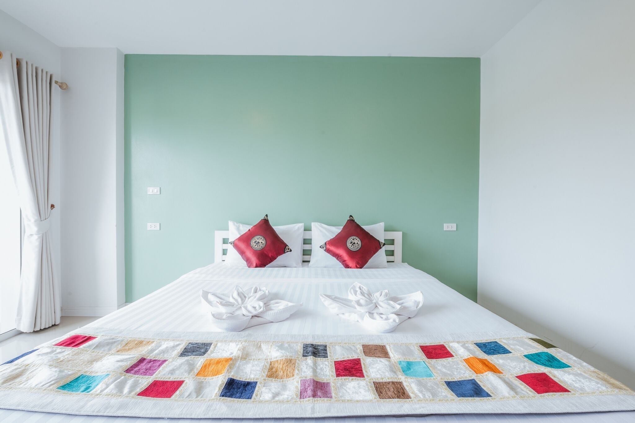 Cama bem arrumada com colcha colorida, duas almofadas vermelhas e cabeceira branca em quarto com parede verde claro.