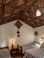 Quarto com telhado de palha, duas camas com roupa de cama estampada, uma cadeira de madeira e uma luminária fixada na parede.