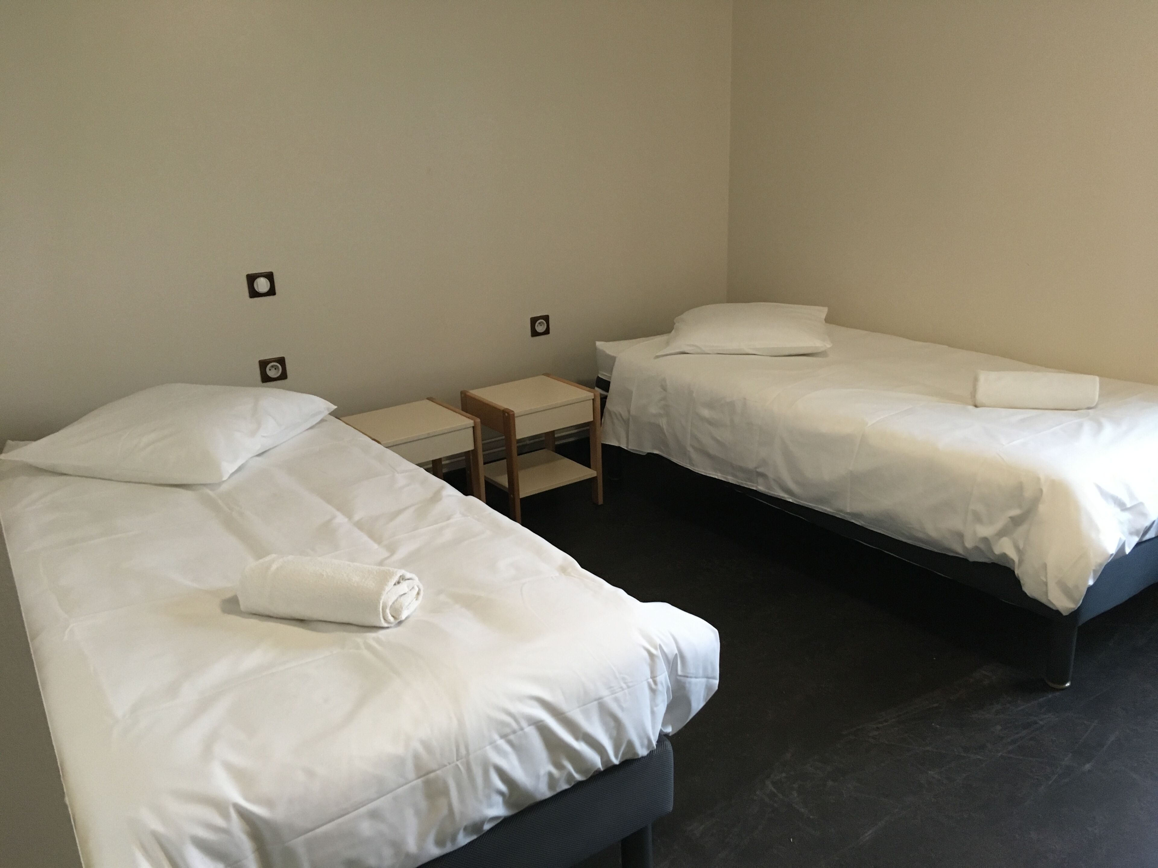 Quarto de hotel com duas camas de solteiro, uma mesinha de cabeceira e um criado-mudo.
