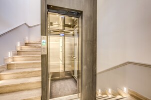 Um elevador moderno com porta de vidro, ao lado de uma escadaria com degraus de mármore e corrimão.