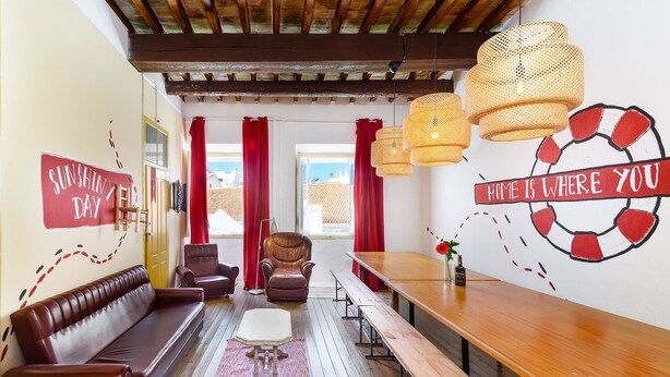 Uma sala de estar acolhedora com um sofá, cadeiras, uma mesa de madeira e um banco. Há cortinas vermelhas, um abajur de vime e uma parede com o letreiro 'Sunshine Day' e a obra de arte 'Home is where you'.