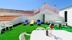 Terraço no topo do prédio com grama sintética verde, mesas e cadeiras brancas, e poltronas de lounge azuis e amarelas.