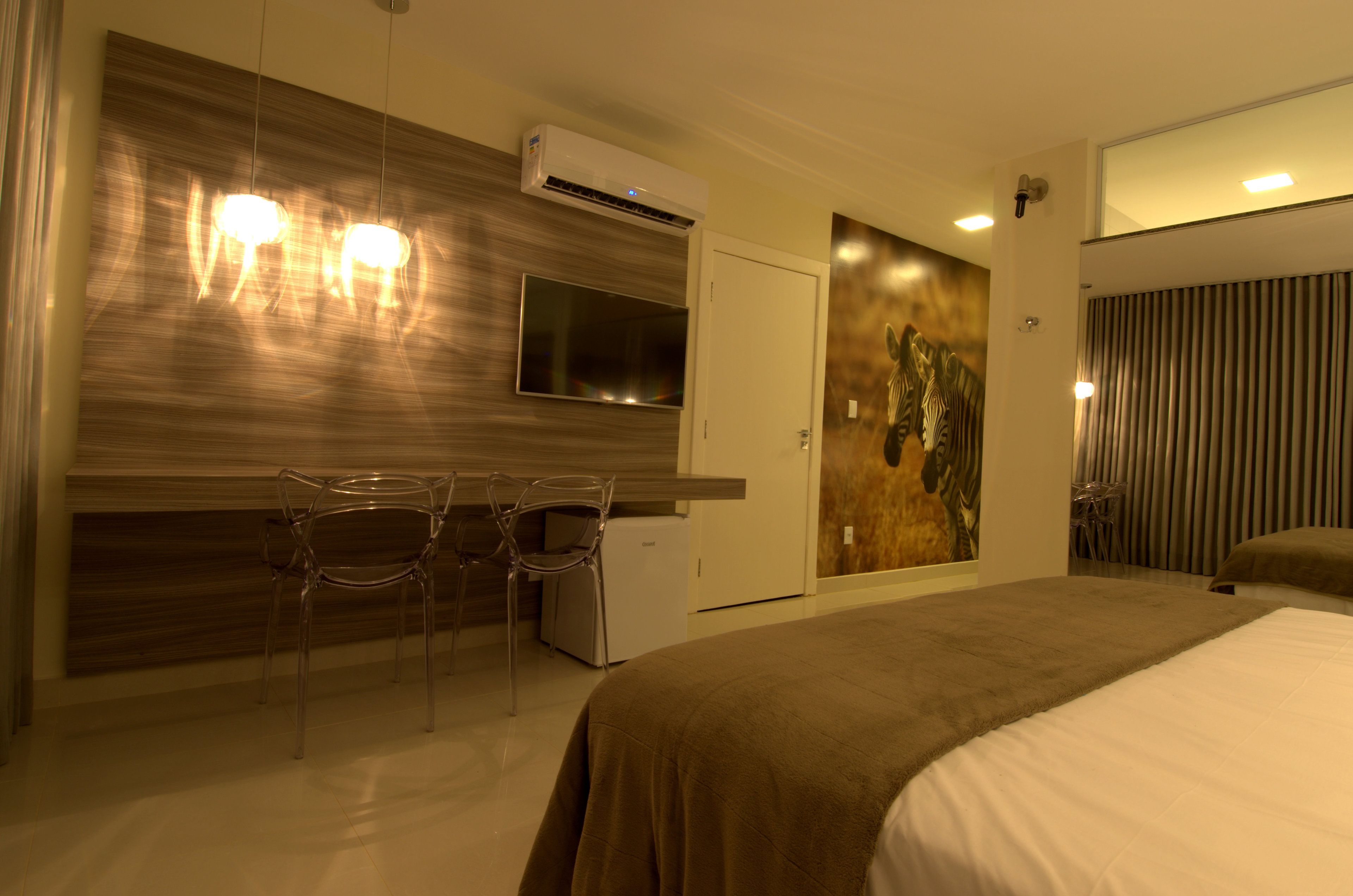 Quarto de hotel com cama, televisão na parede, pequeno frigorífico e área de estar com cadeiras.