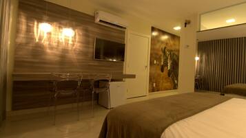 Quarto de hotel com cama, televisão na parede, pequeno frigorífico e área de estar com cadeiras.