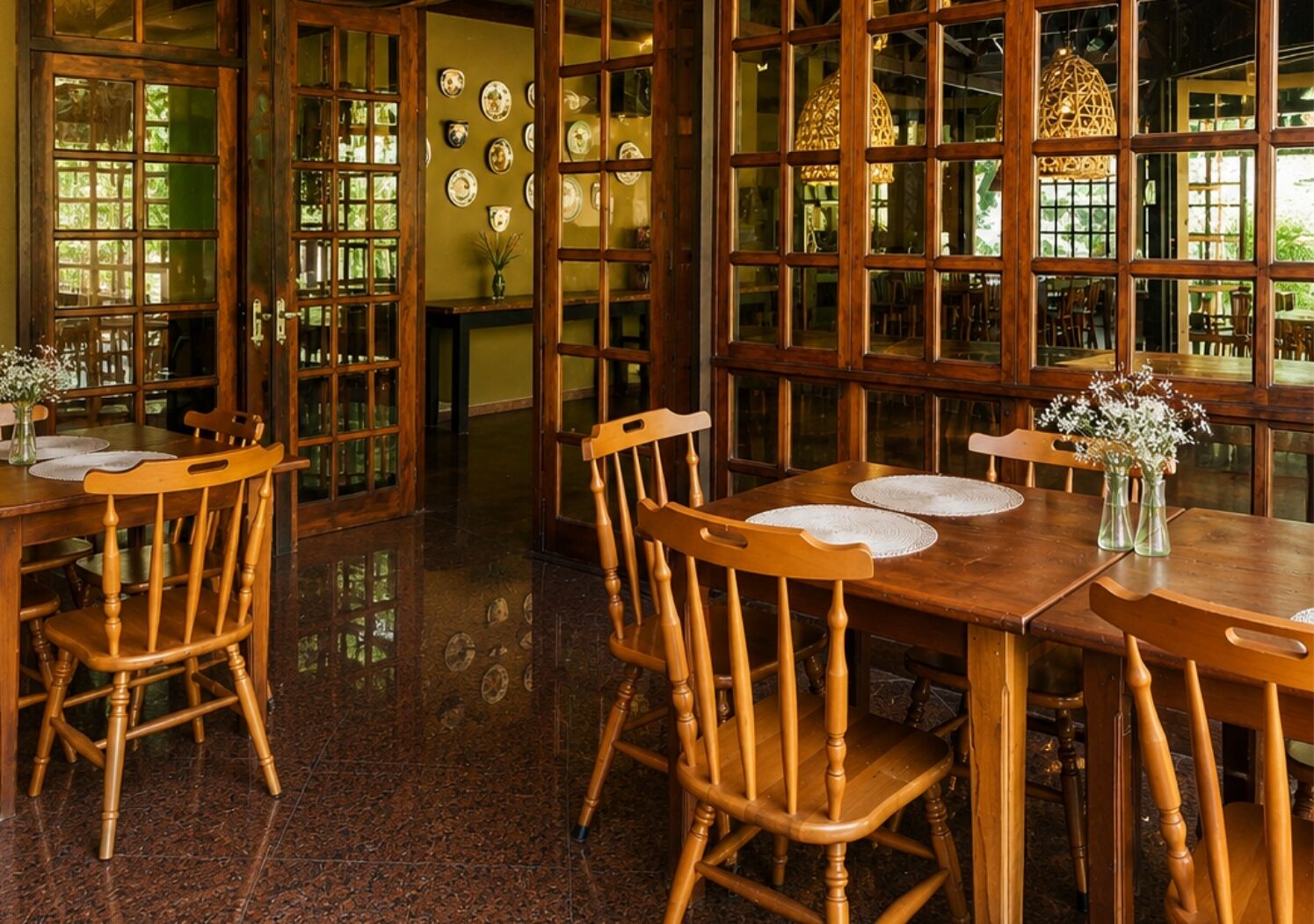 Uma sala de jantar com mesas e cadeiras de madeira, parede decorada com pratos e vista através de grandes janelas.