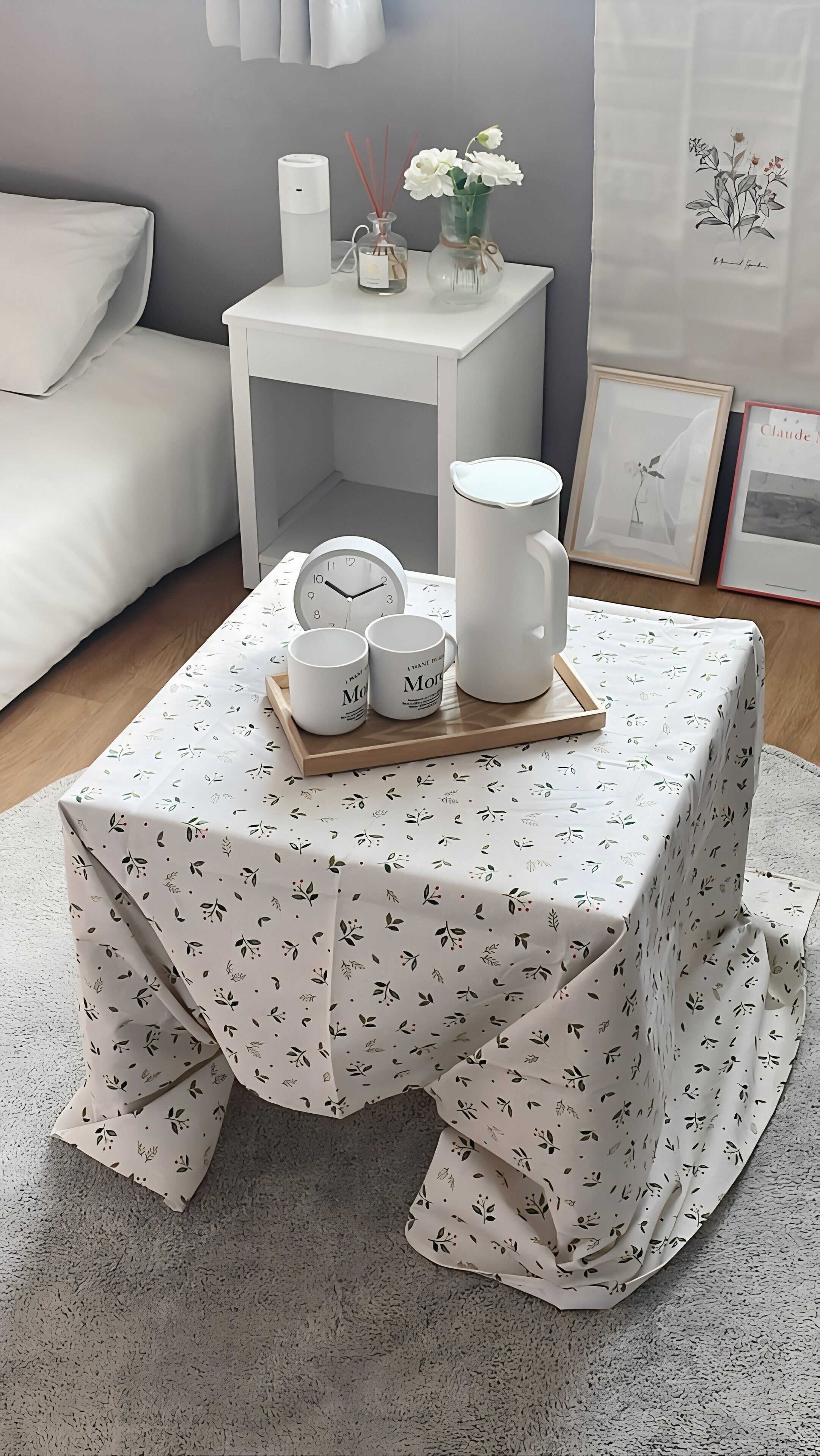 Uma mesa bem arrumada com toalha de mesa florida, um relógio branco, duas canecas, um jarro branco e um pequeno vaso com flores.