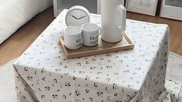 Uma mesa bem arrumada com toalha de mesa florida, um relógio branco, duas canecas, um jarro branco e um pequeno vaso com flores.