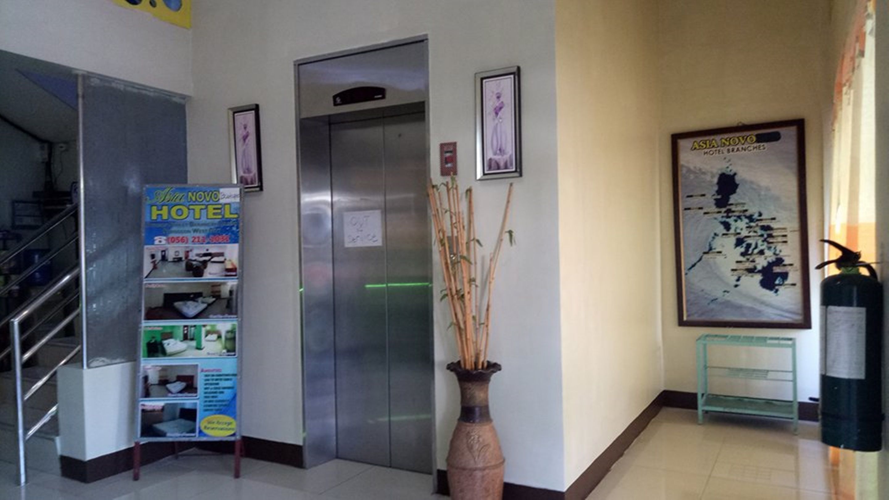 Um saguão de hotel com elevador, balcão de recepção, uma planta de bambu e um mapa emoldurado na parede.