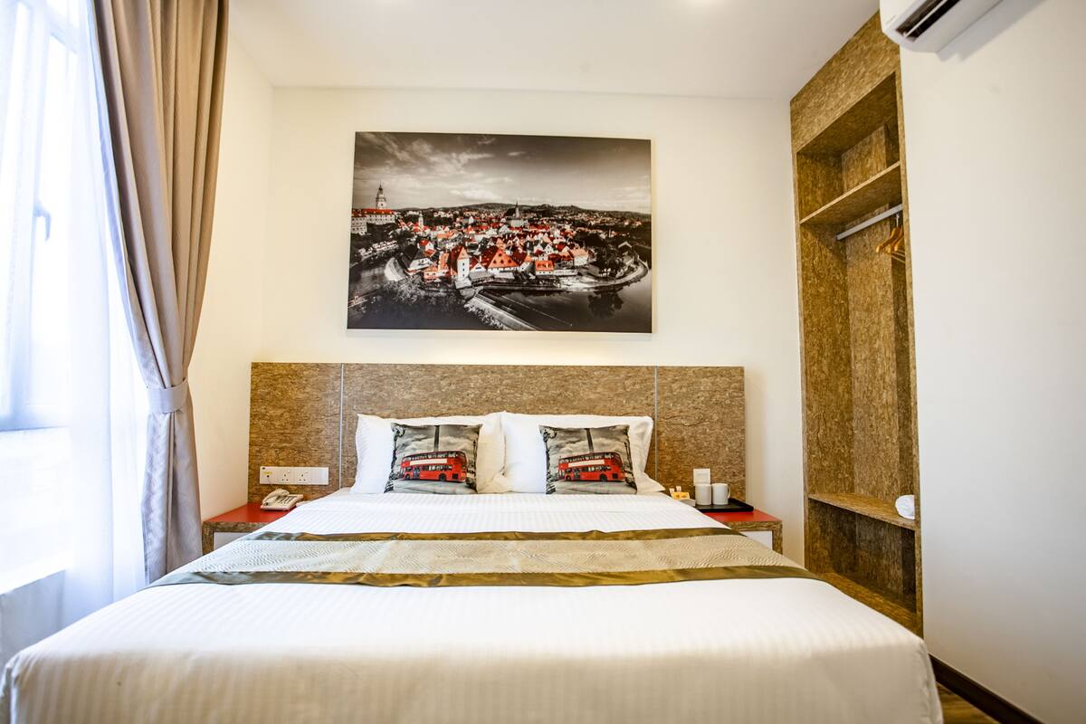 Quarto de hotel com uma cama grande, uma mesinha de cabeceira, ar condicionado fixado na parede e uma pintura com vista para a cidade sobre a cama.