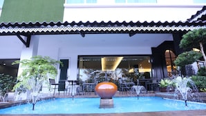 Uma piscina com uma fonte central, rodeada por plantas em vasos e mobiliário de exterior.