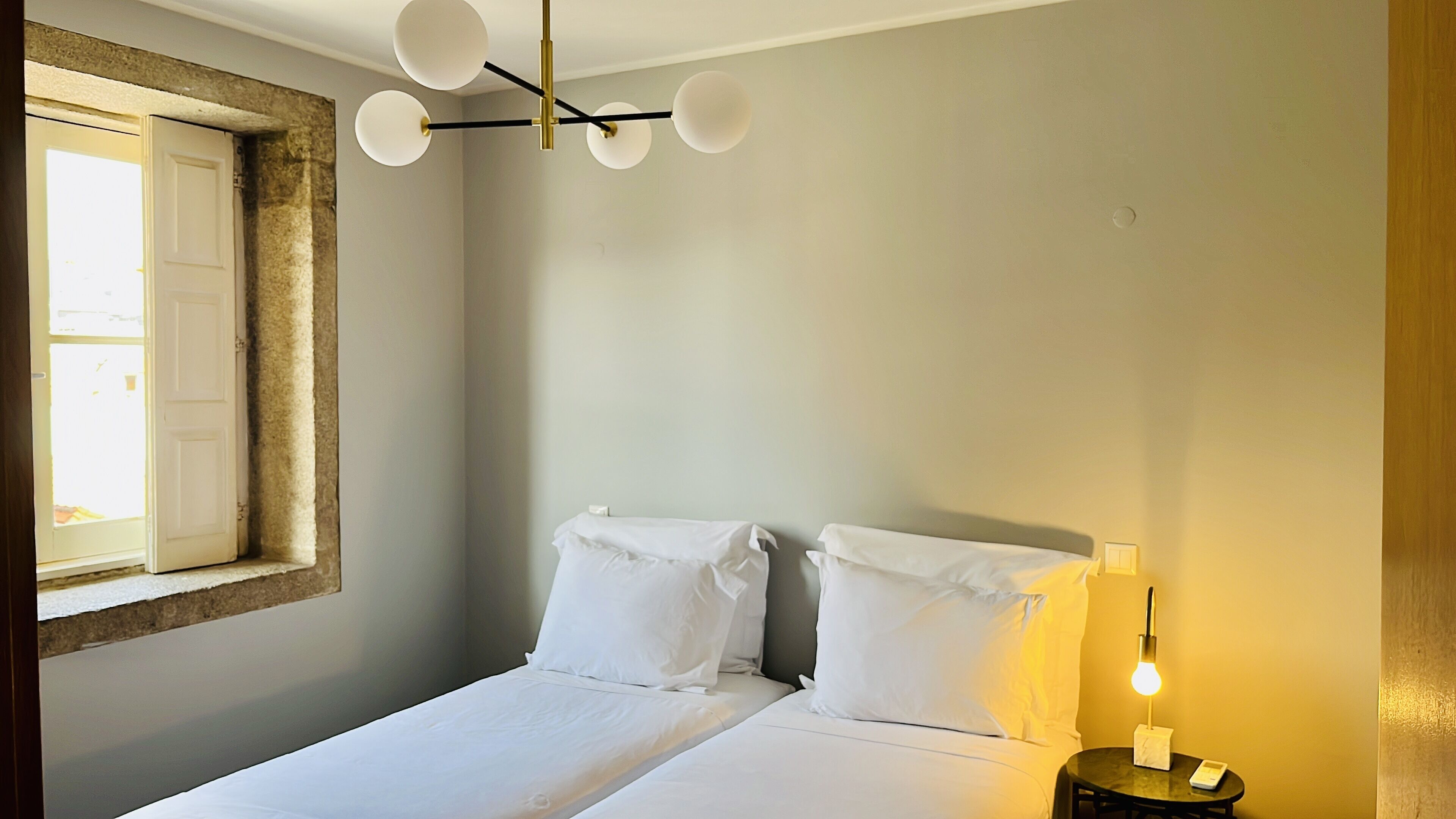 Quarto com cama, duas almofadas, mesa de cabeceira com abajur, janela e luminária no teto.