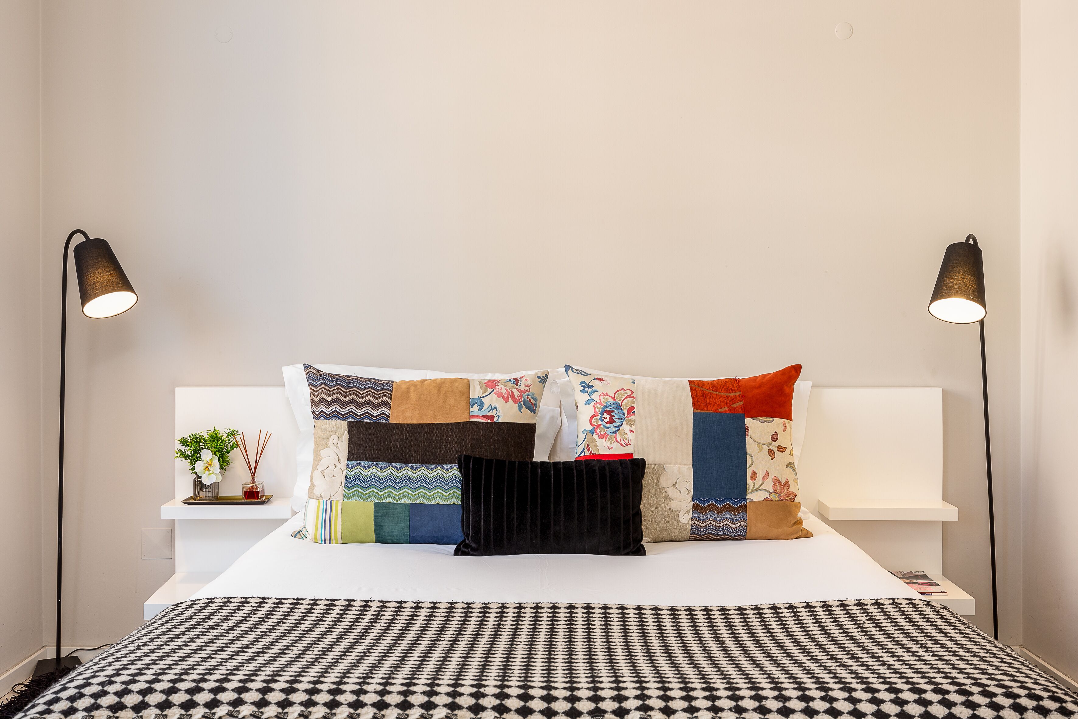 Cama bem arrumada com colcha estampada, vários travesseiros e duas luminárias de cabeceira.