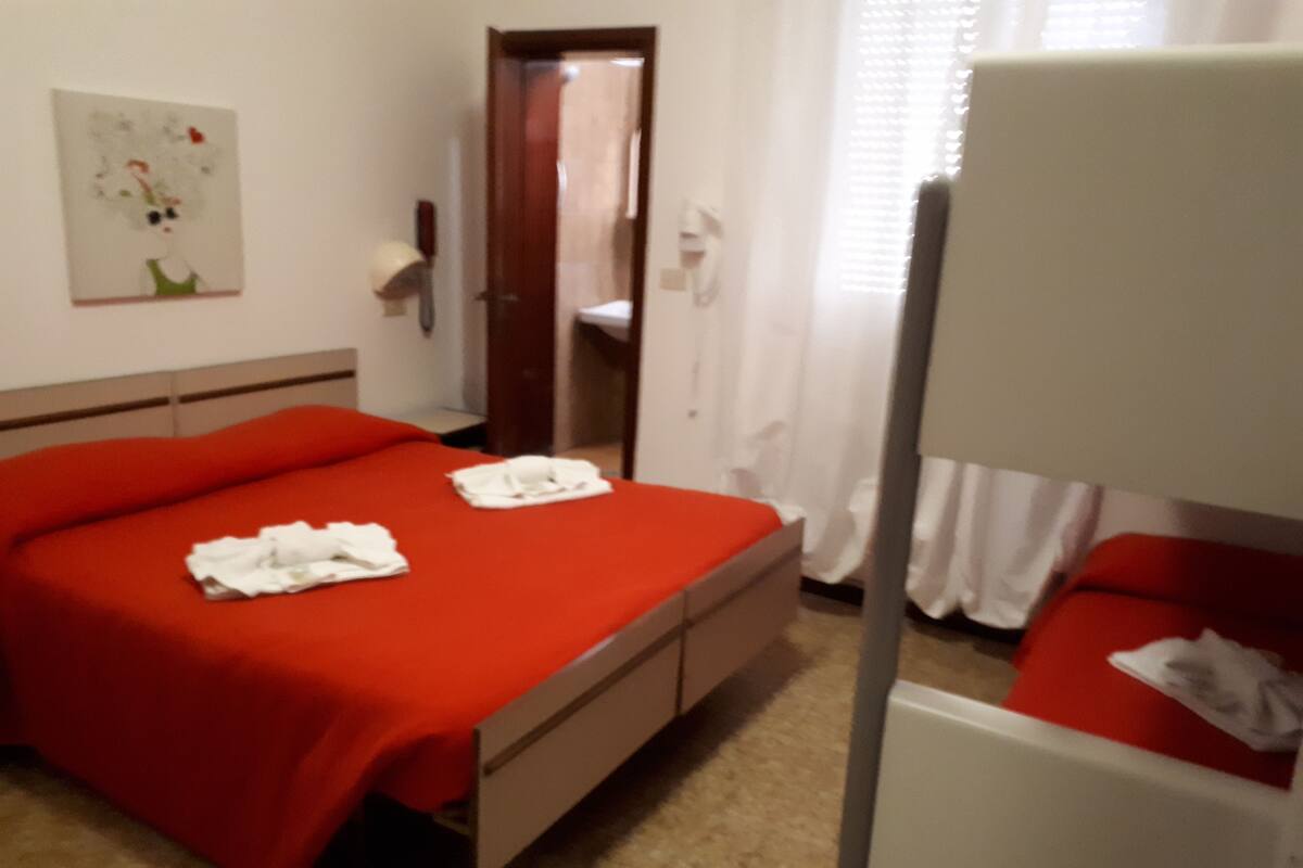 Quarto de hotel com colcha vermelha, mesa de cabeceira branca e uma cadeira pequena branca.