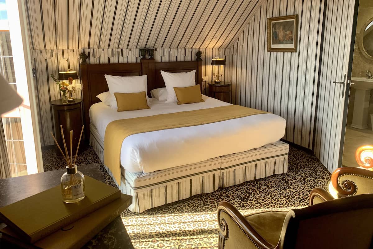 Quarto de hotel com uma cama grande, duas mesas de cabeceira, um criado-mudo, uma cadeira, um espelho e um tapete estampado.