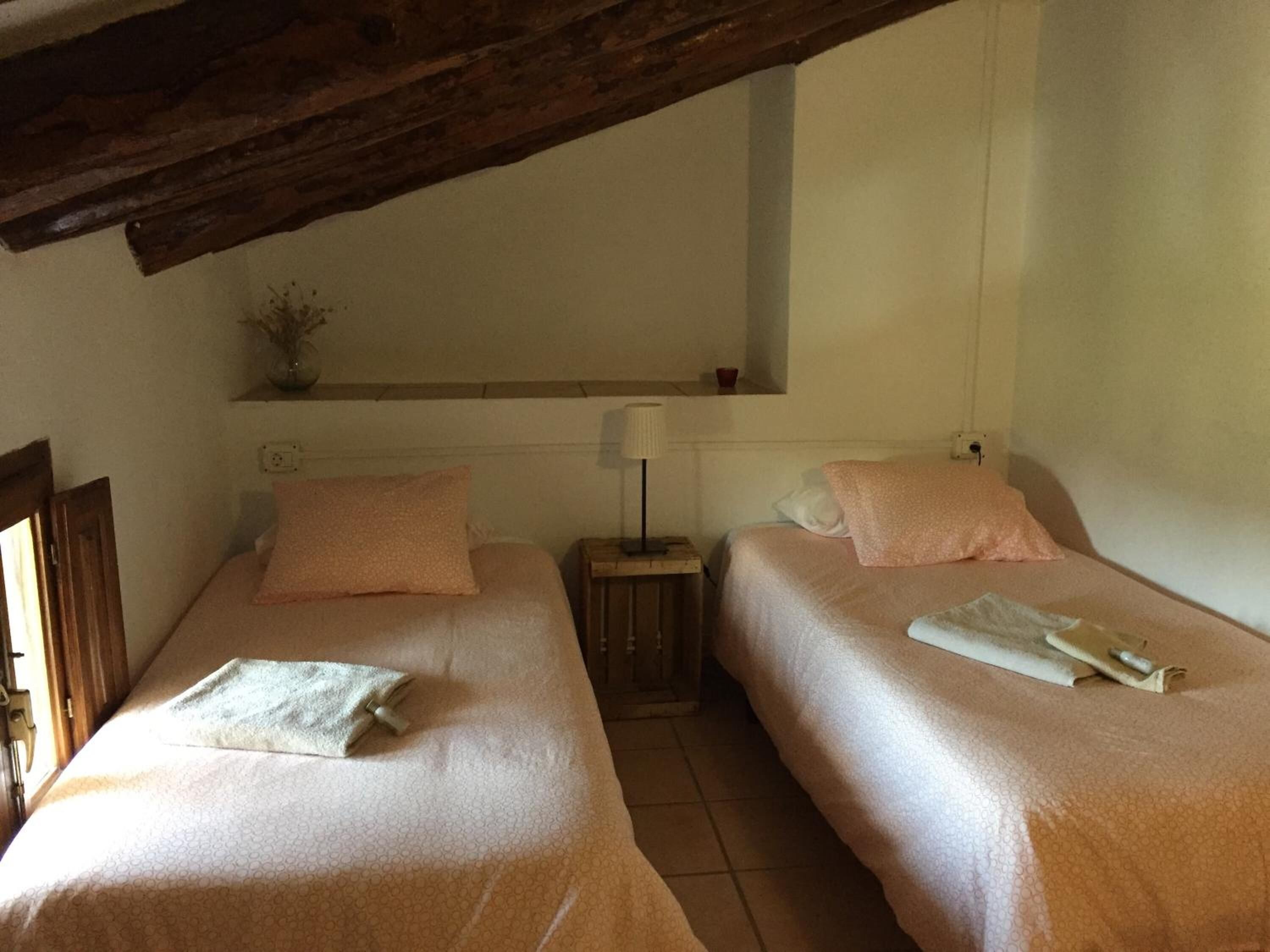 Quarto com duas camas, cada uma com colcha e almofadas cor-de-rosa, uma pequena mesa de cabeceira com abajur e uma prateleira sobre as camas.