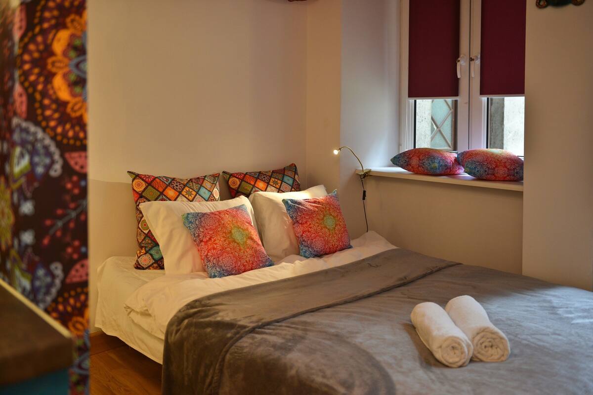 Uma cama bem arrumada com almofadas coloridas, uma janela com cortinas vermelhas e um abajur.