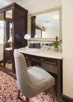 Quarto de hotel com uma escrivaninha de madeira, uma cadeira, um espelho, telefone, abajur, caixa de lenços, planta em vaso e carpete estampado.
