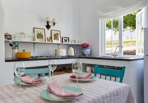 Uma sala de jantar com mesa posta para dois, janela com vista para vinhedo e bancada de cozinha com diversos objetos.