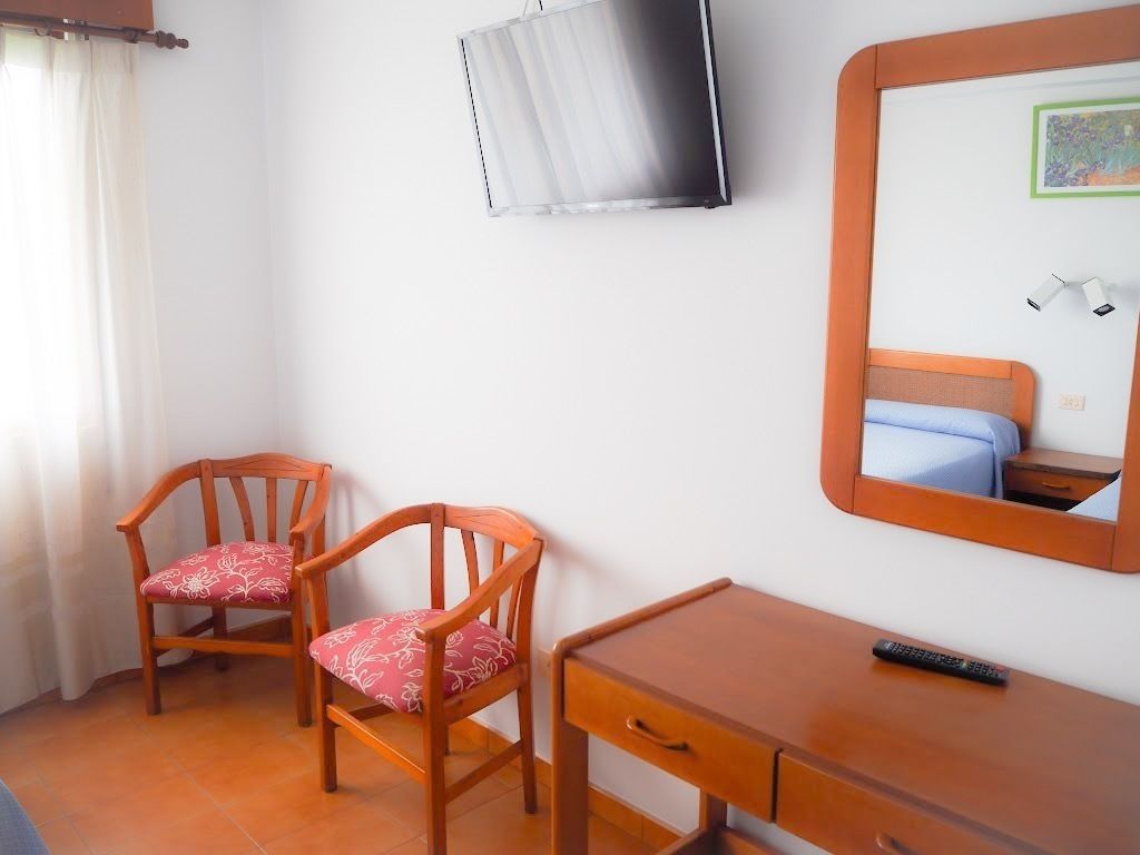 Quarto com duas cadeiras de madeira com almofadas estampadas, uma escrivaninha de madeira com gaveta, uma televisão presa na parede e um espelho refletindo uma cama e uma mesinha de cabeceira.
