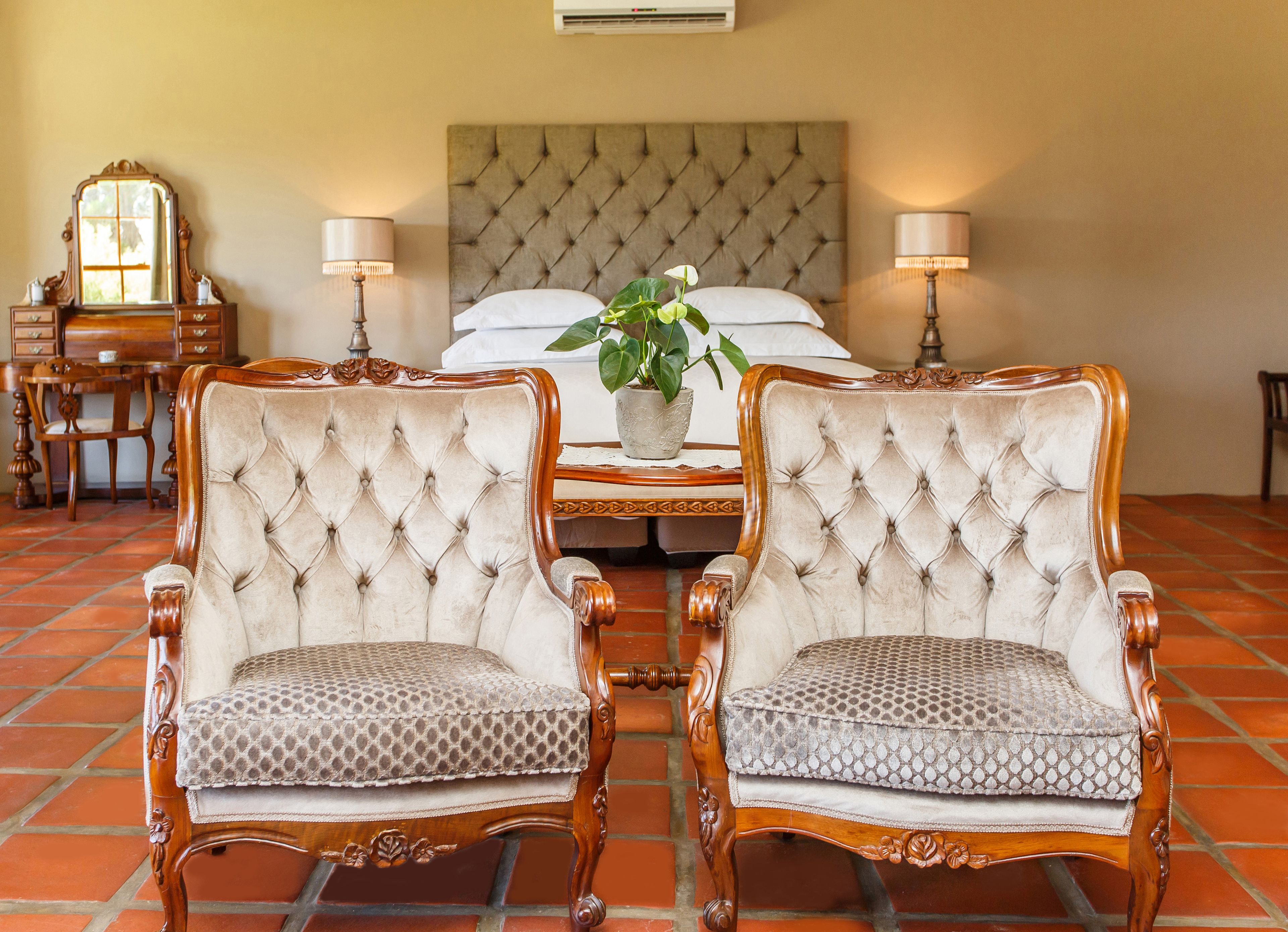 Quarto de hotel com uma cama, duas poltronas estofadas com almofadas estampadas, uma mesinha de cabeceira com abajur, cômoda e um vaso de planta sobre a cama.